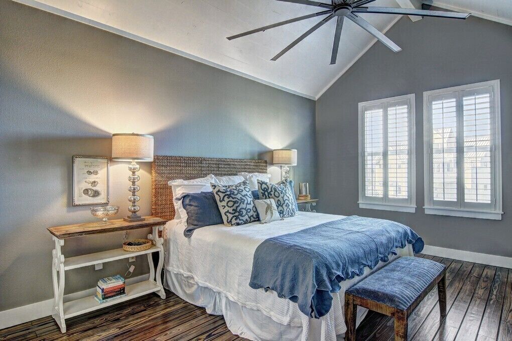 Quarto com teto inclinado, cama com colcha branca e azul, duas luminárias de cabeceira, mesa de cabeceira com abajur, quadro emoldurado e janela com persianas.