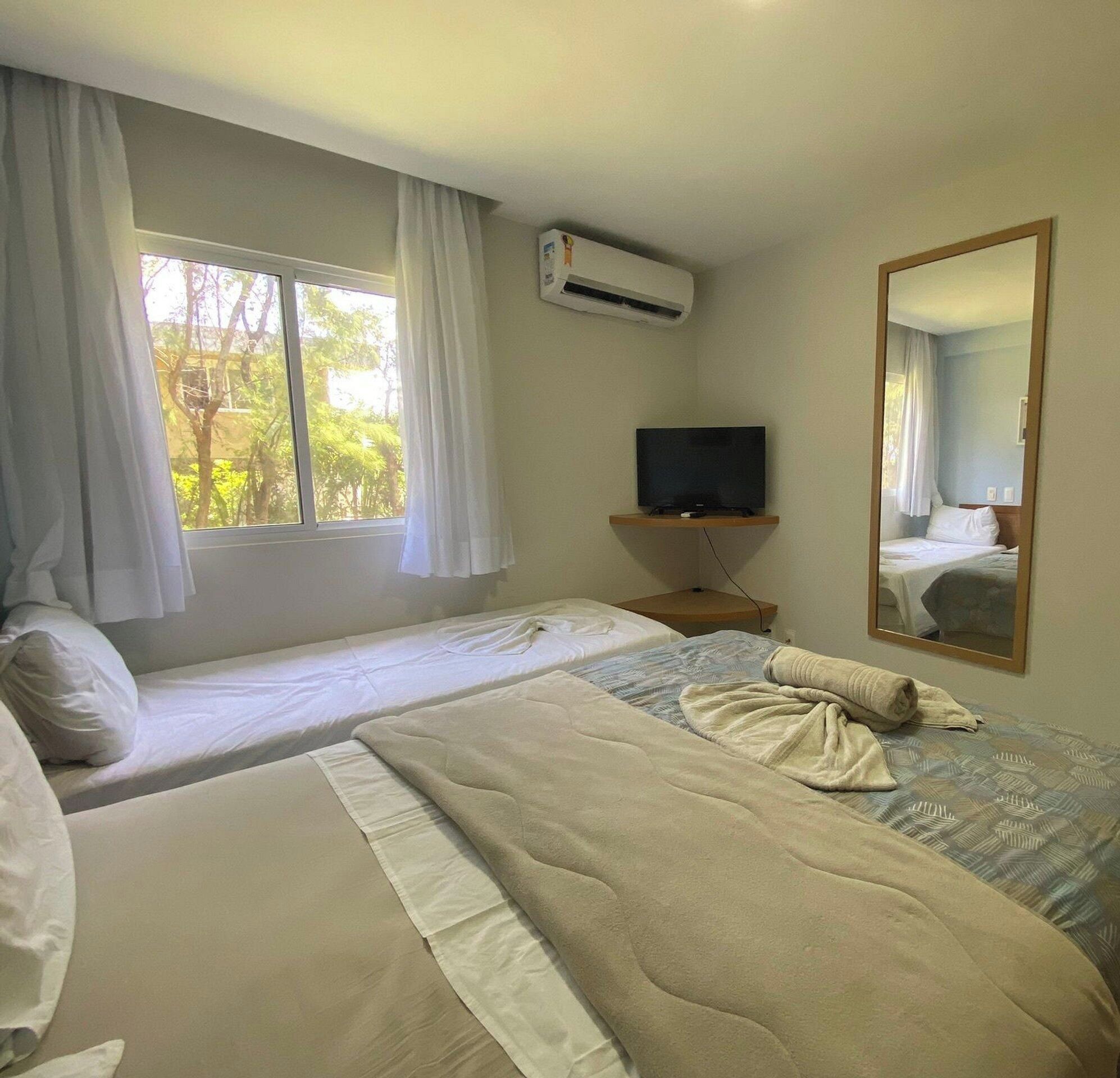 Quarto de hotel com duas camas, televisão em um suporte, aparelho de ar condicionado e um espelho grande na parede.