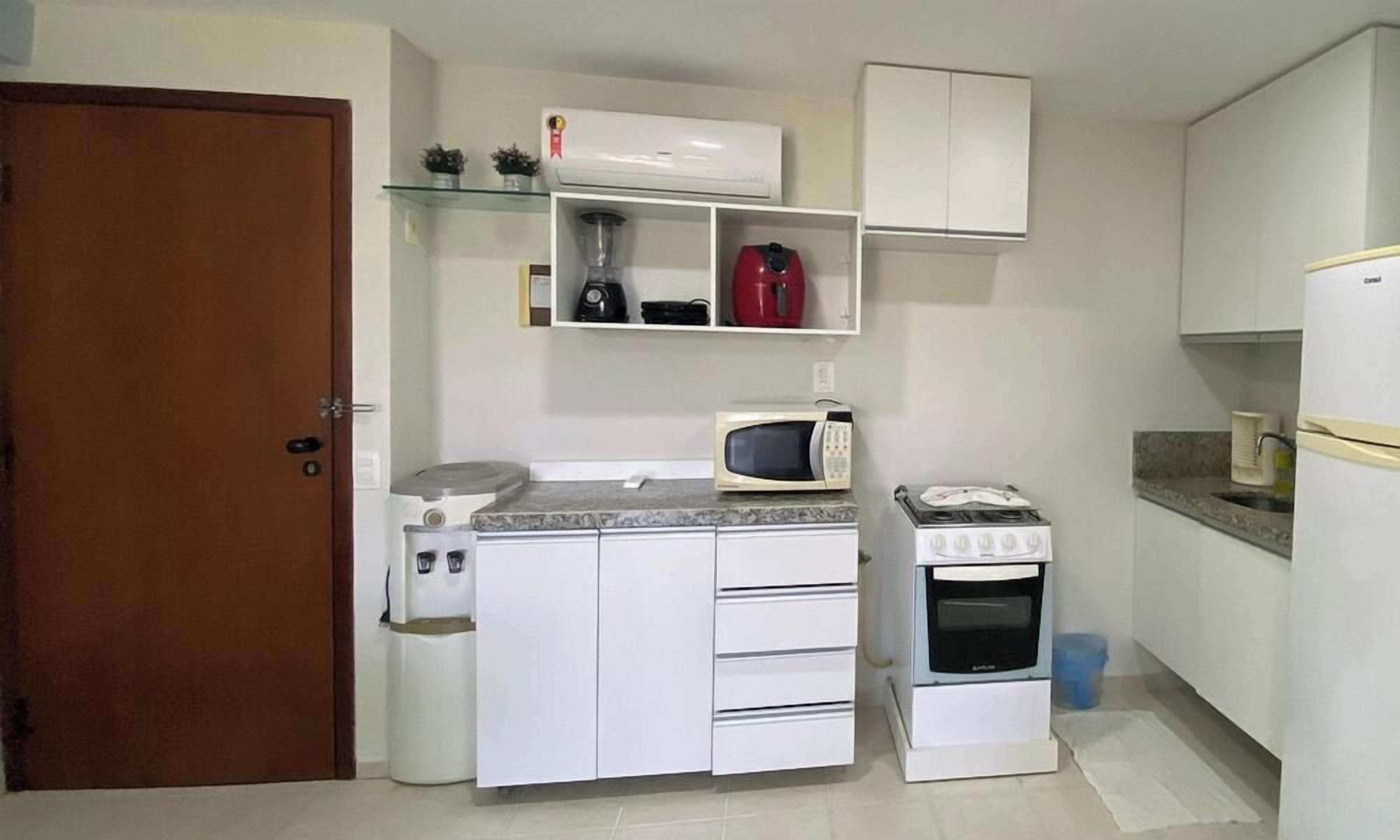 Uma cozinha compacta com armários brancos, micro-ondas, fogão, dispenser de água e uma pequena prateleira com um liquidificador e um aparelho vermelho.