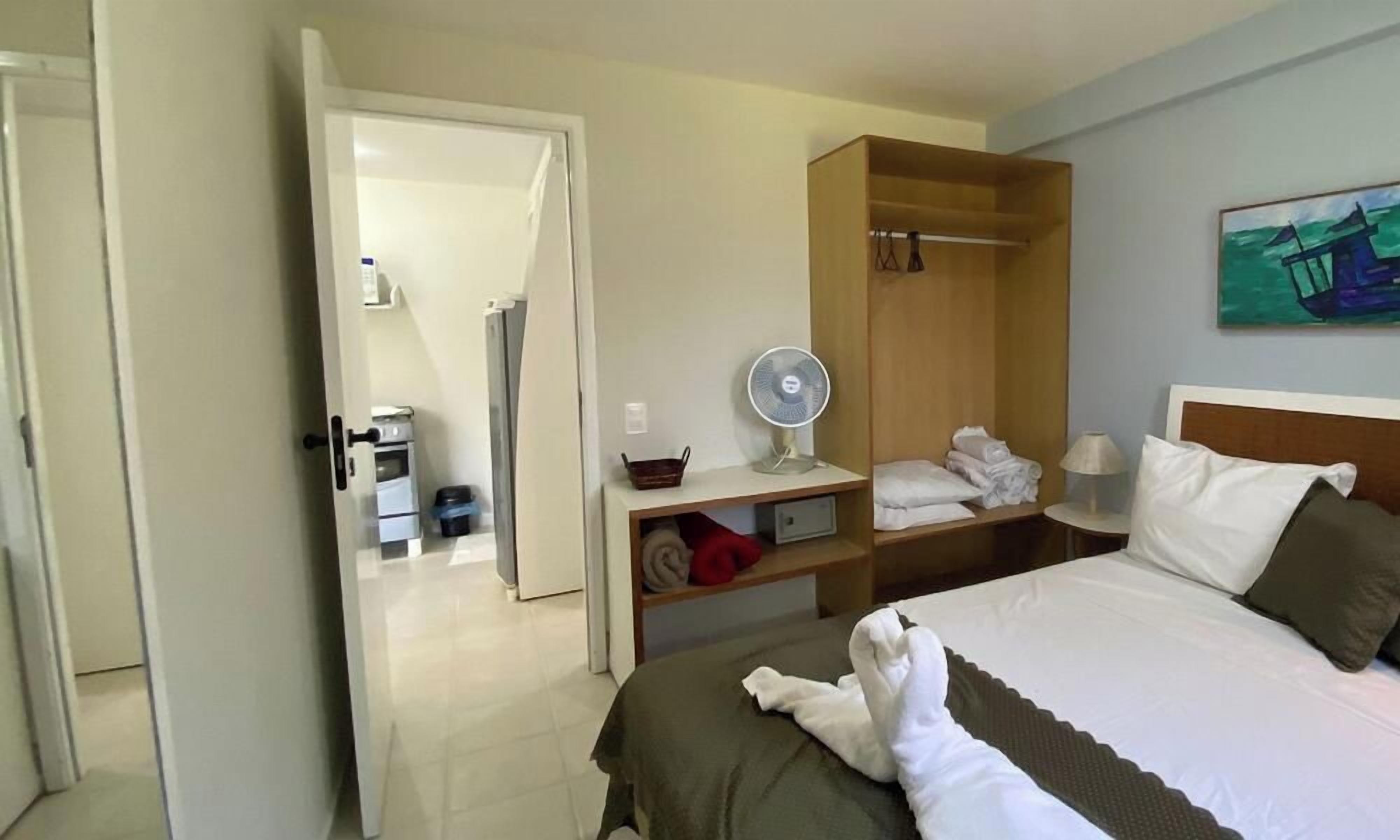 Quarto de hotel com cama, mesa de cabeceira com luminária, ventilador, prateleira com toalhas e um quadro na parede.