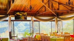 Uma área de refeições interna com mesas e cadeiras, grandes janelas e estrutura de teto em madeira.