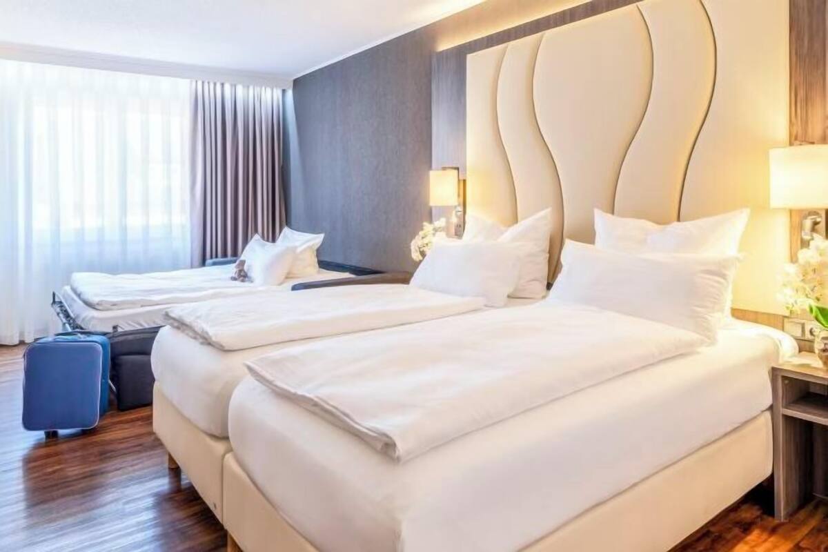 Quarto de hotel com duas camas, uma janela grande com cortinas, piso de madeira e mesa de cabeceira com abajur.