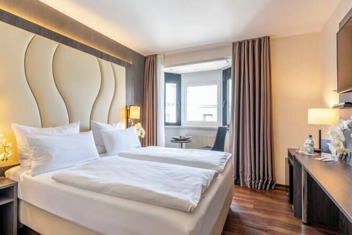 Quarto de hotel com cama grande, mesas de cabeceira, escrivaninha e vista para o exterior.