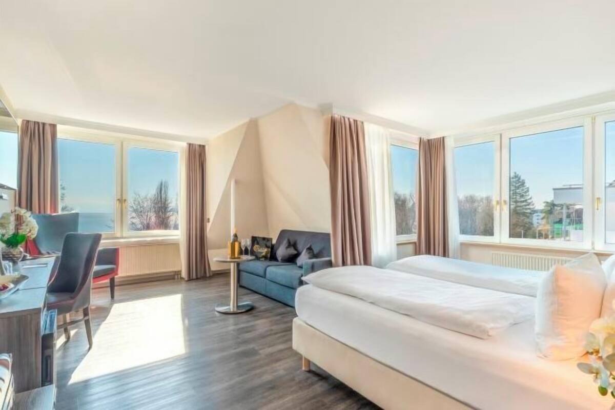 Quarto de hotel com cama, escrivaninha, cadeira, sofá e grandes janelas.