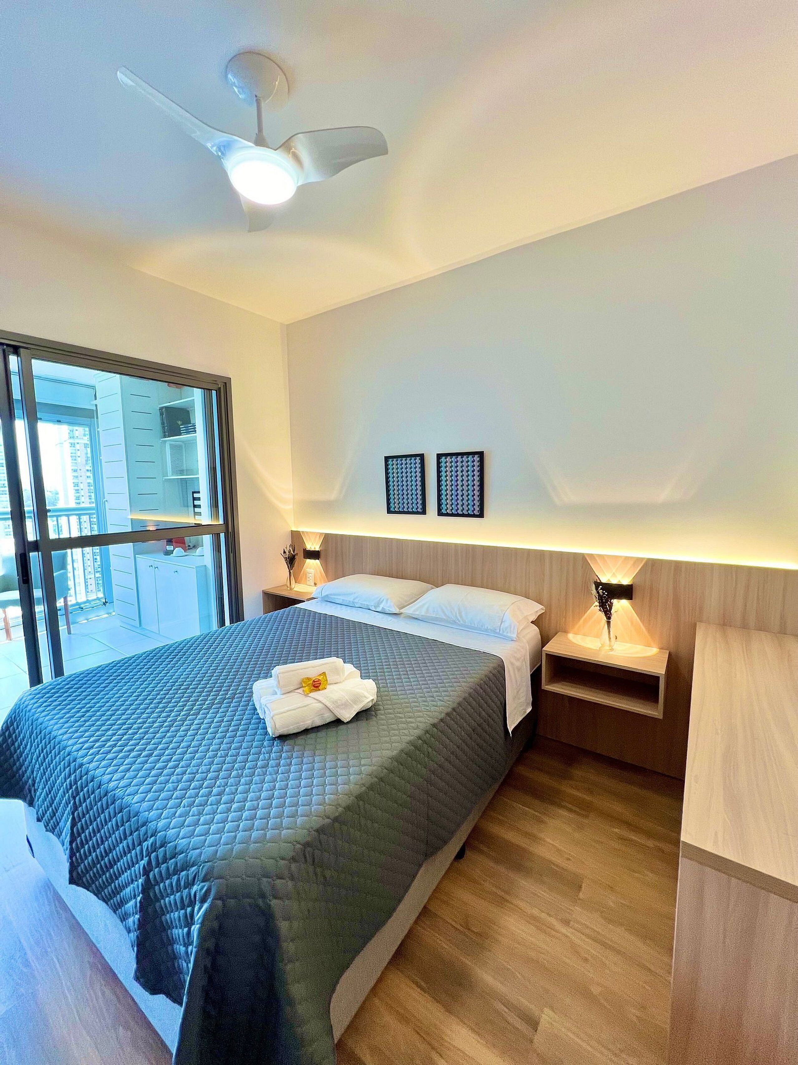 Quarto de hotel moderno com uma cama grande, um ventilador de teto, mesa de cabeceira com abajur e vista para a varanda.