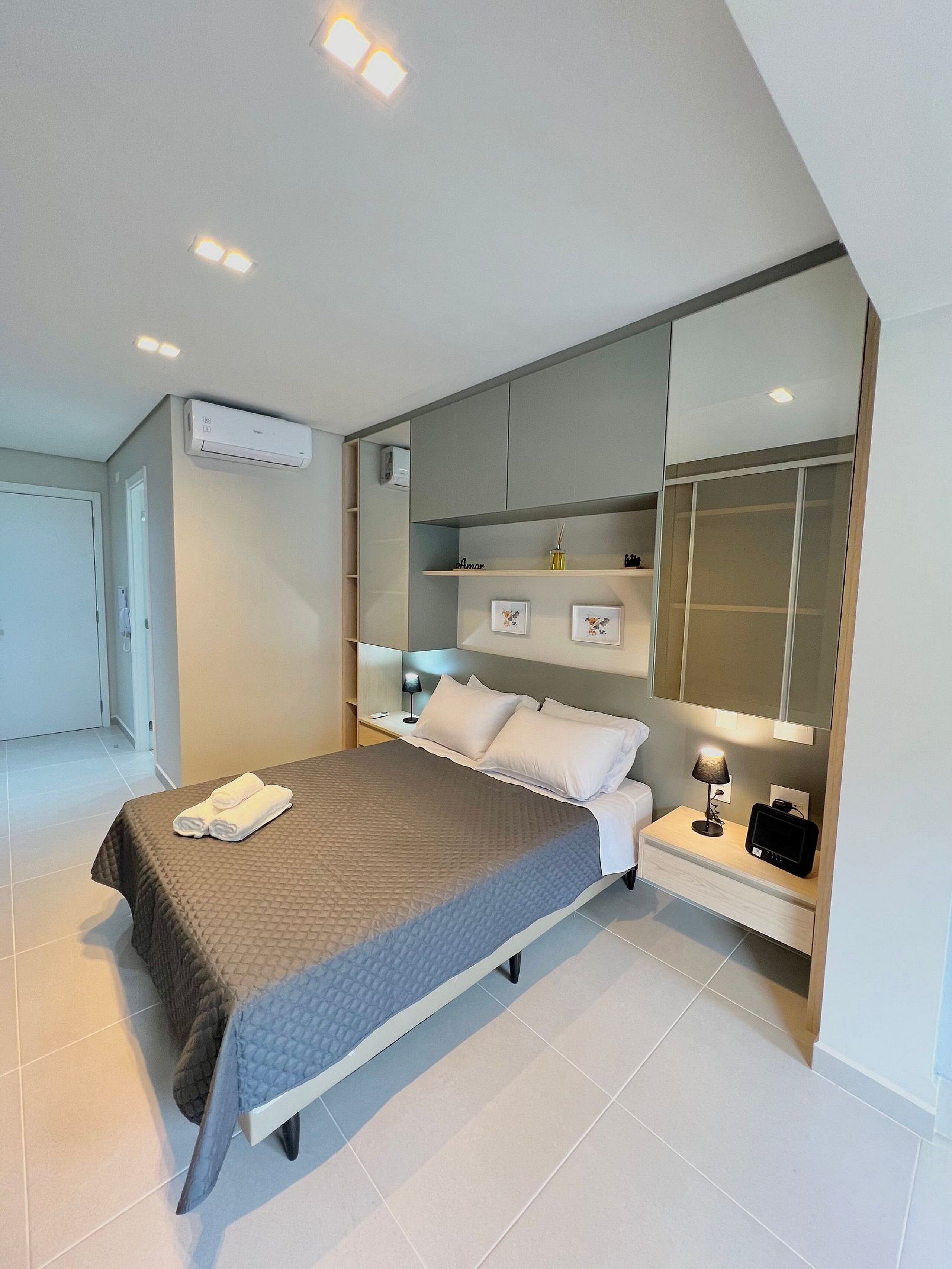 Um quarto moderno com cama, mesas de cabeceira, um criado-mudo, ar-condicionado fixado na parede e armários embutidos.