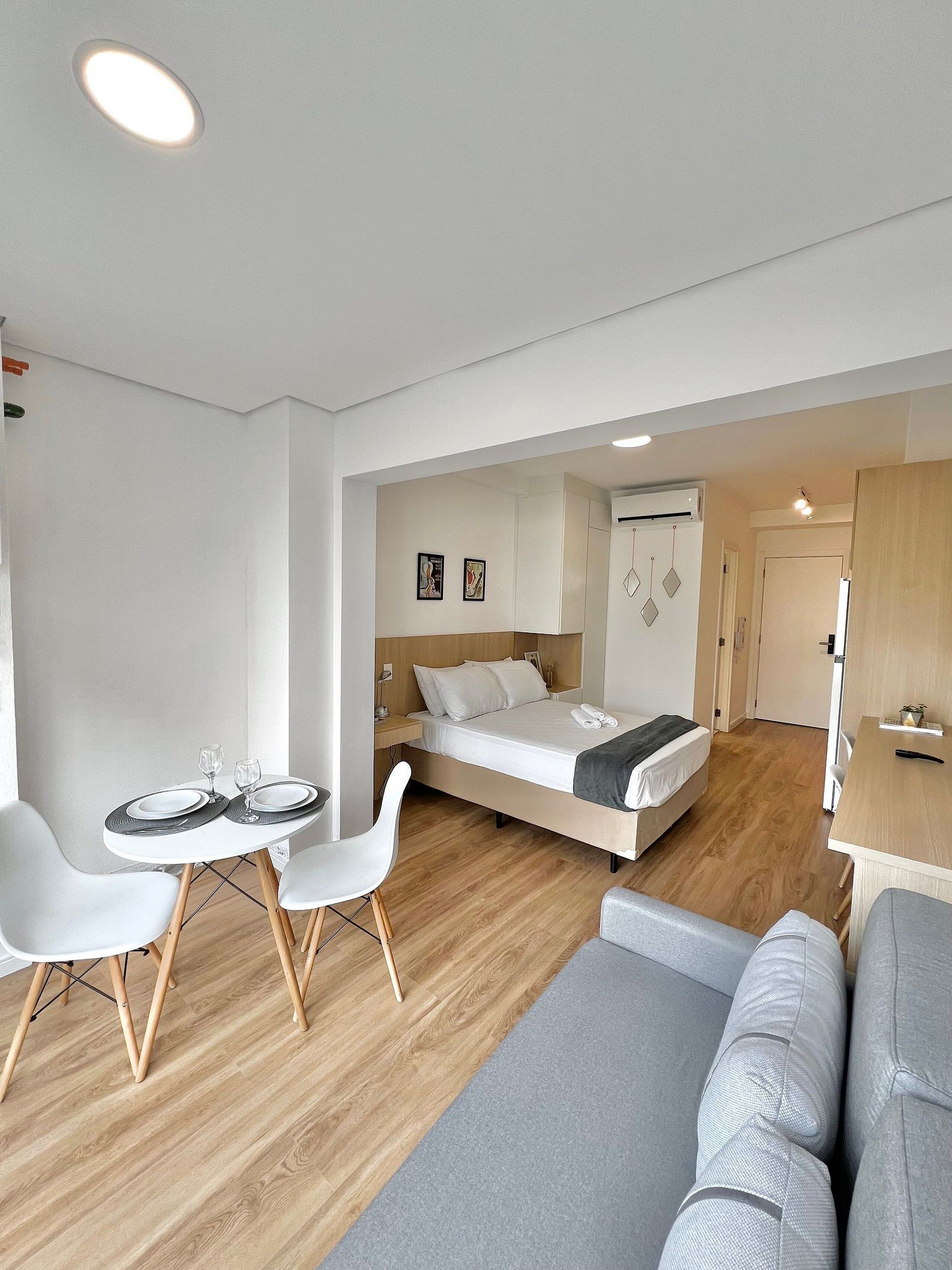 Quarto de hotel moderno com cama, mesa de jantar com cadeiras, sofá e cozinha compacta.