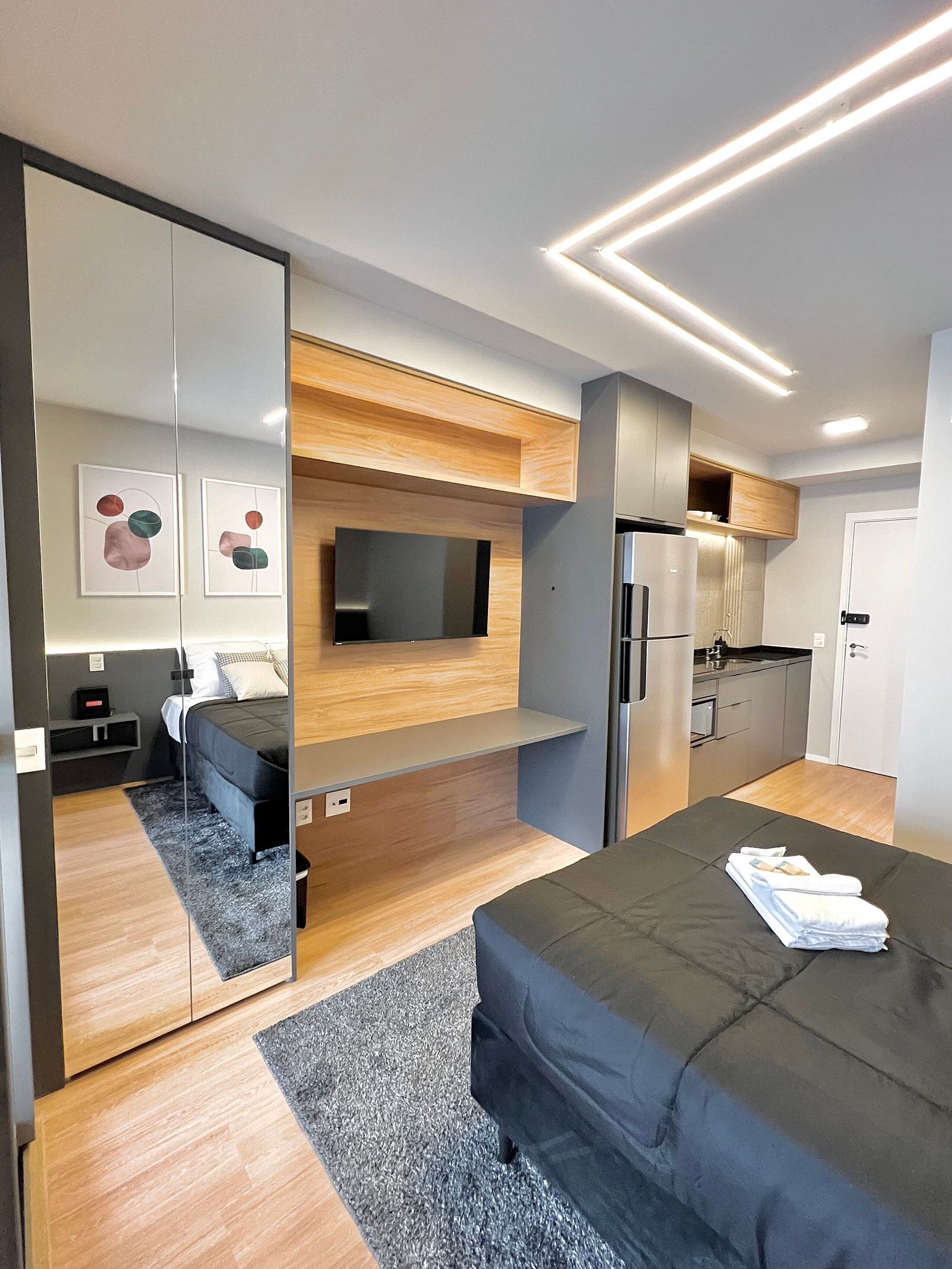 Quarto de hotel moderno com cama, televisão na parede, área de cozinha e banheiro.