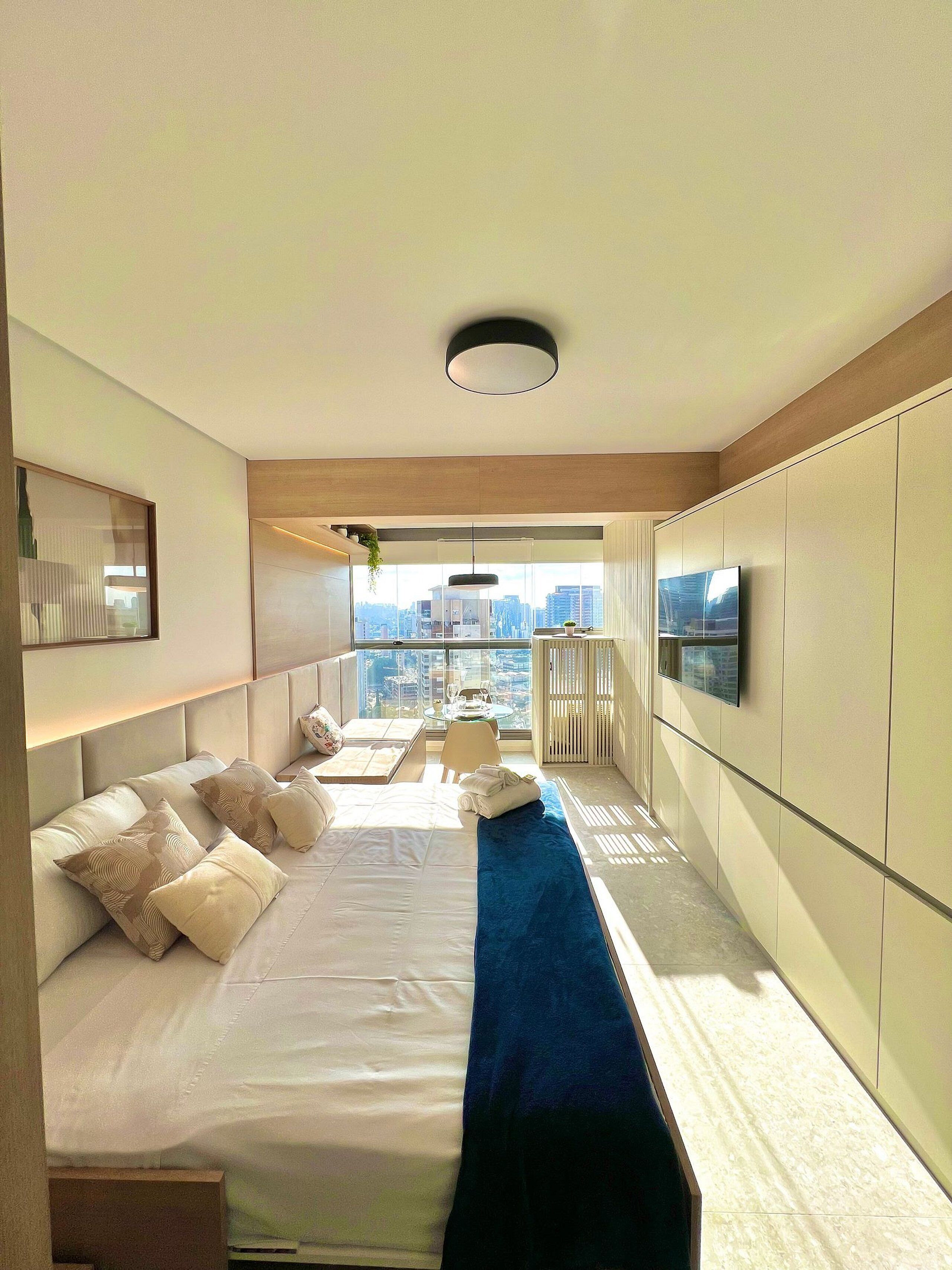 Quarto de hotel moderno com uma cama grande, televisão, varanda com vista para a cidade e uma janela ampla.