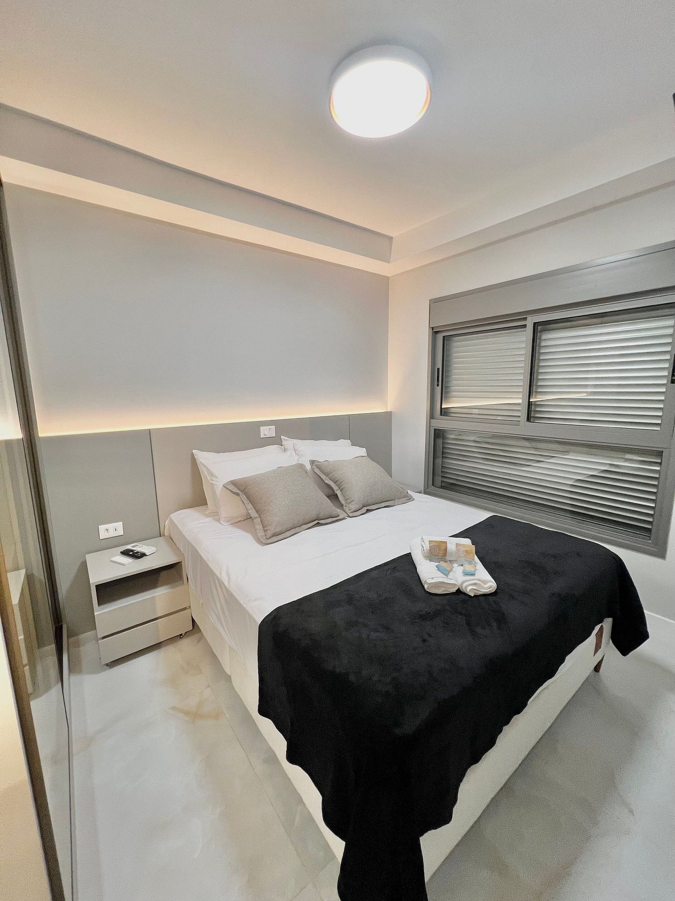 Quarto de hotel moderno com cama, mesa de cabeceira e janela com persianas.