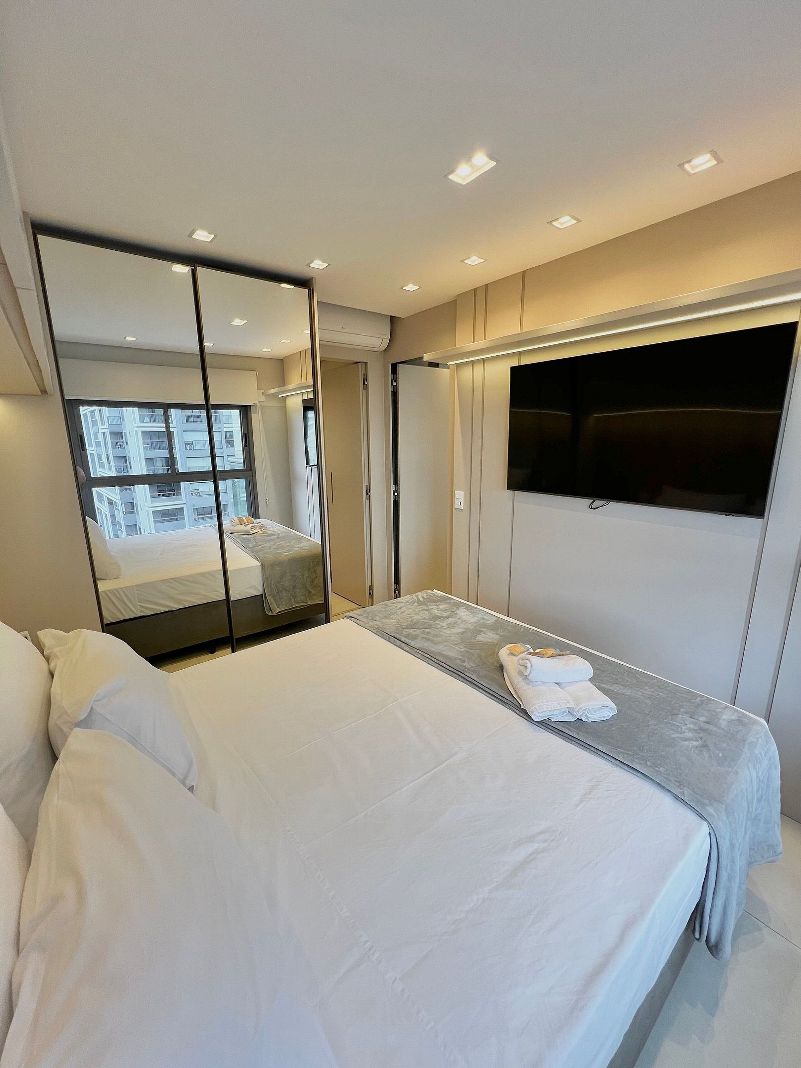 Quarto de hotel moderno com uma cama grande, televisão fixada na parede e uma janela ampla com vista para a cidade.