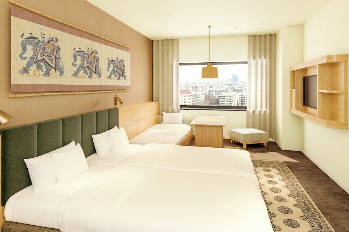 Quarto de hotel com uma cama grande, uma cama menor, uma escrivaninha, uma televisão, uma janela com vista para a cidade e uma tapeçaria decorativa.