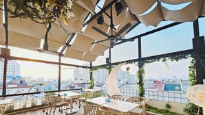 Um terraço no telhado com piso de madeira, mesas brancas, cadeiras de vime e um grande ventilador.