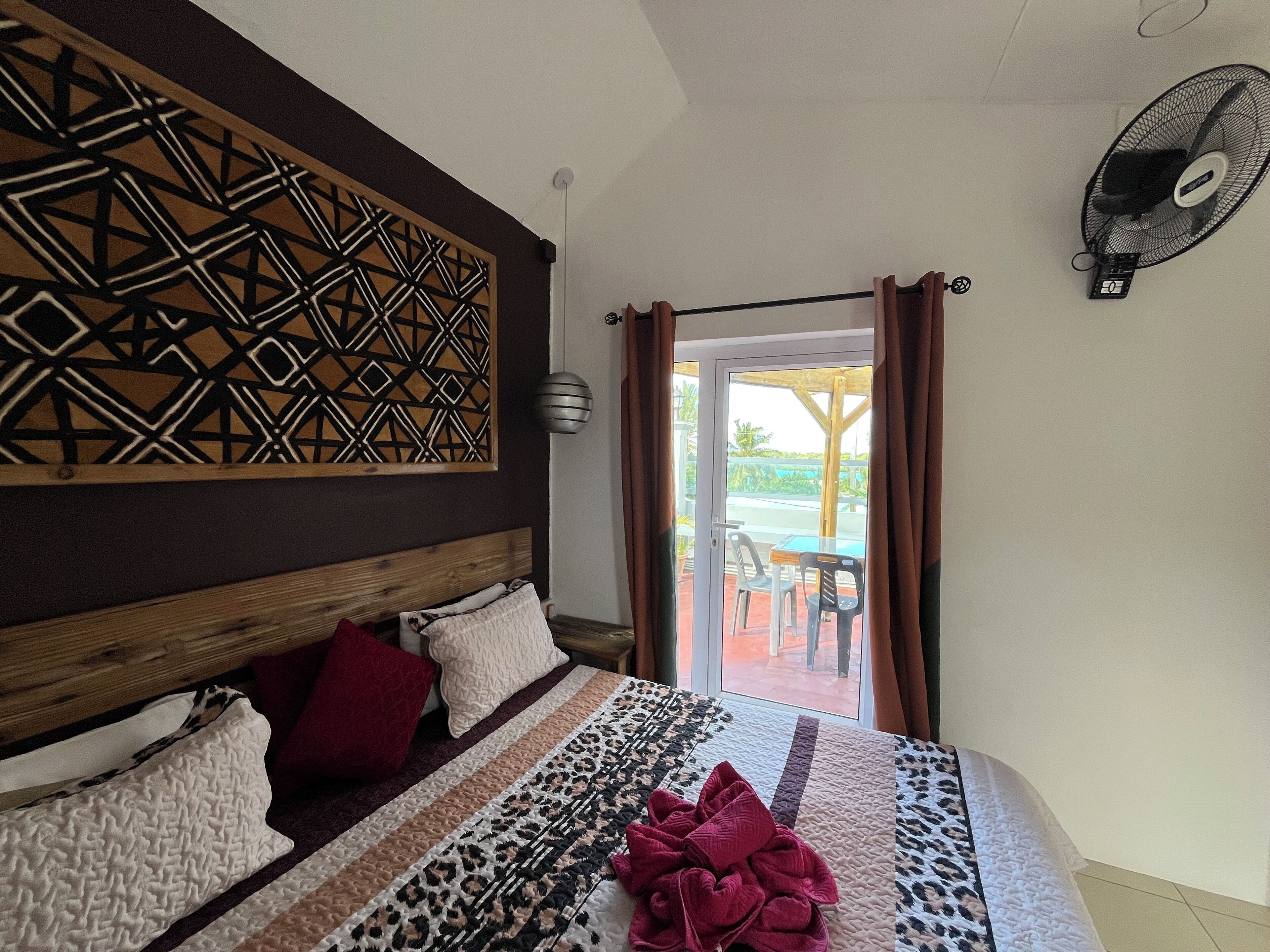 Quarto com cabeceira estampada, cama com várias almofadas, um ventilador e vista para um pátio externo.