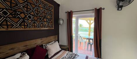 Quarto com cabeceira estampada, cama com várias almofadas, um ventilador e vista para um pátio externo.