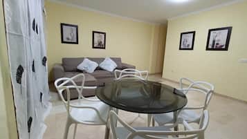 Uma área de refeições com mesa de vidro cercada por cadeiras brancas, um sofá bege com almofadas e quadros nas paredes.