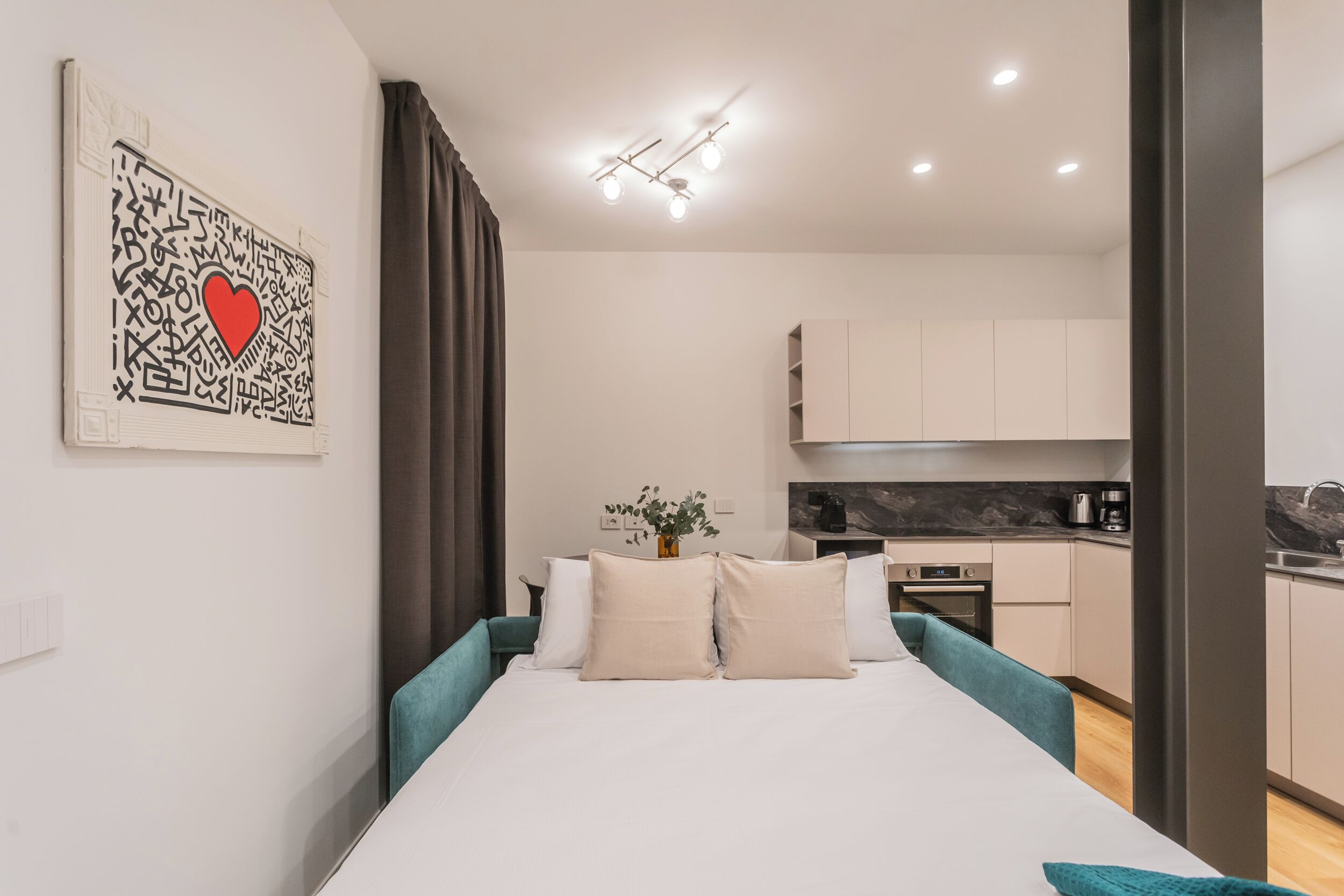 Quarto de hotel moderno com uma cama grande, pequena área de cozinha e um objeto decorativo na parede.