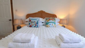 Cama bem arrumada com almofadas brancas e cabeceira, duas luminárias de cabeceira e toalhas dobradas sobre a cama.