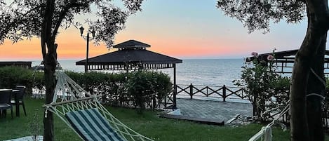 Uma rede sob uma árvore, um gazebo e vista para o mar ao pôr do sol.