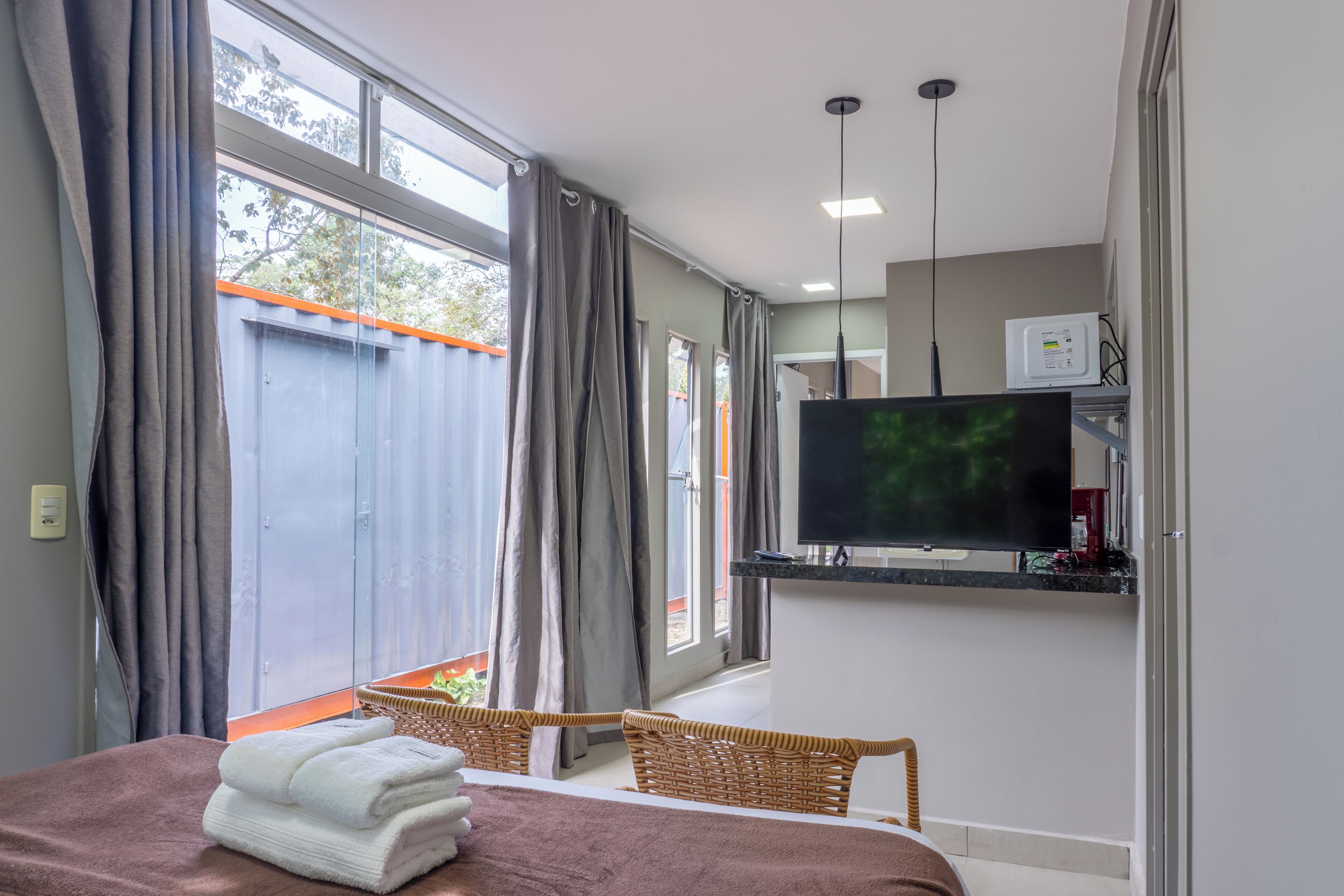 Quarto de hotel com cama, televisão, uma pequena mesa com toalhas, cadeiras de vime e vista para o exterior.