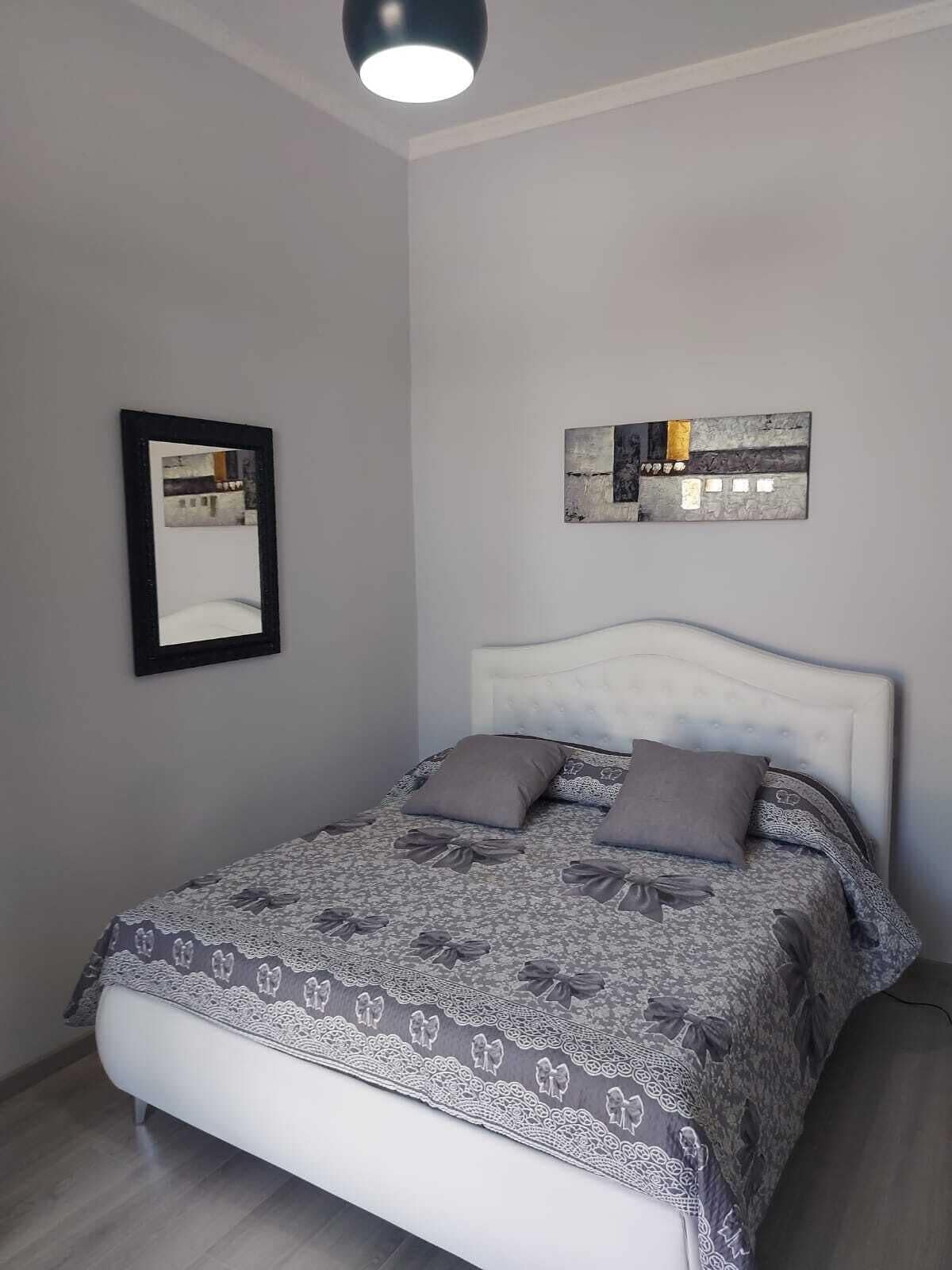 Quarto com cama branca, colcha de linhas cinzas, duas almofadas, espelho e uma obra de arte na parede.