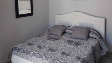 Quarto com cama branca, colcha de linhas cinzas, duas almofadas, espelho e uma obra de arte na parede.