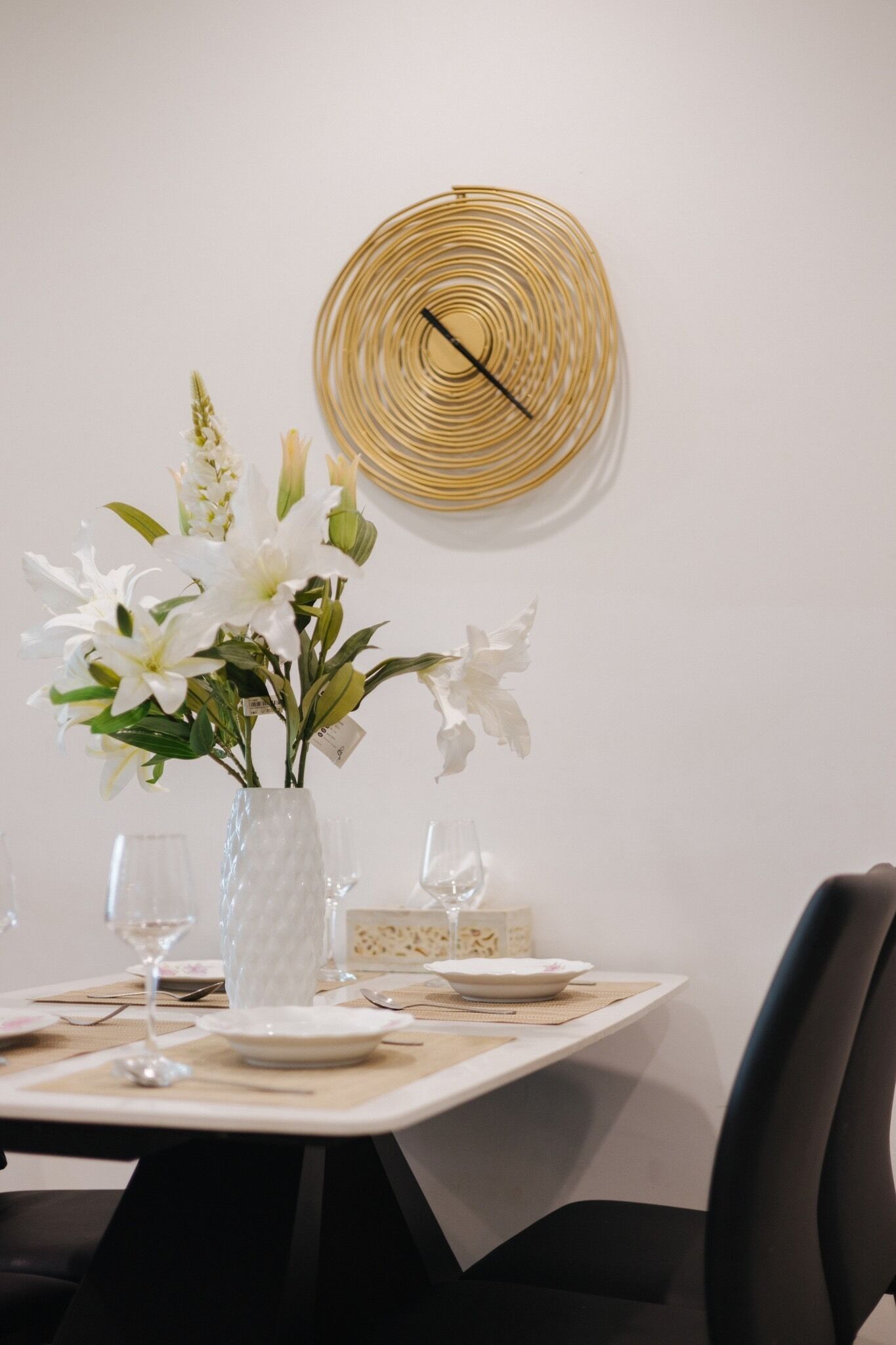 Mesa de jantar posta com pratos brancos, um vaso branco com flores e um enfeite de parede trançado.