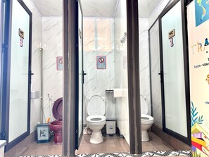 Casa de banho pública com três sanitas, cada uma identificada com um número. Há placas que indicam a proibição de fumar e de lixo. As paredes estão revestidas com um padrão semelhante a mármore.