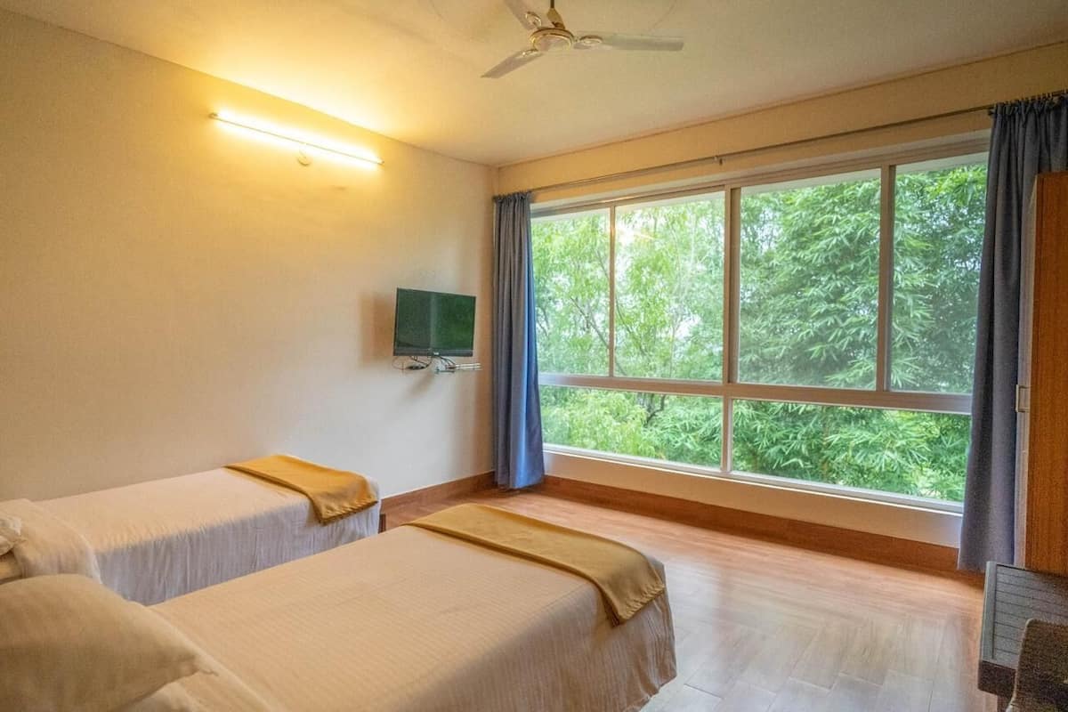 Quarto de hotel com duas camas, ventoinha no teto, televisão e uma janela grande com vista para árvores.