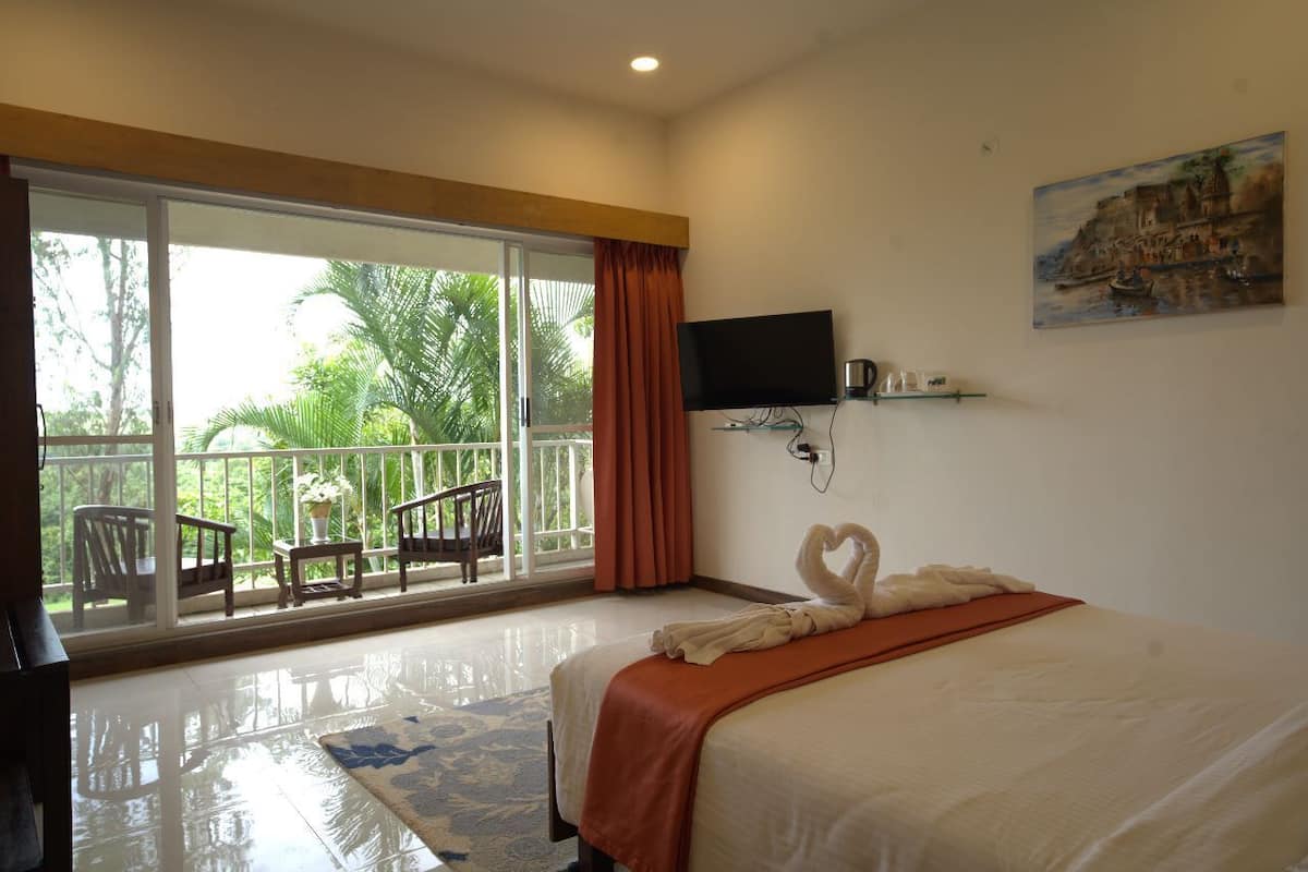 Quarto de hotel com cama, televisão fixada na parede, varanda com cadeiras e um quadro na parede.
