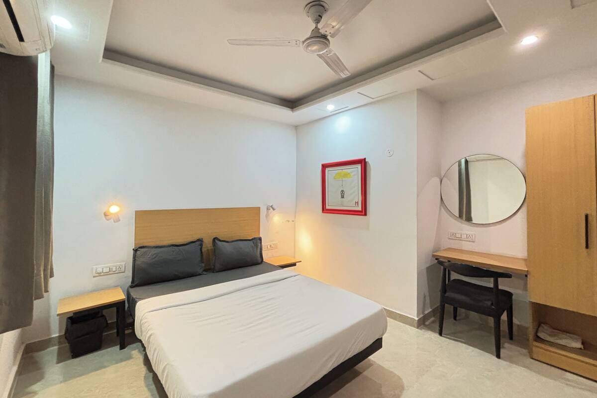 Um quarto com cama, mesas de cabeceira, escrivaninha, cadeira, guarda-roupa, ventoinha no teto e espelho fixado na parede.