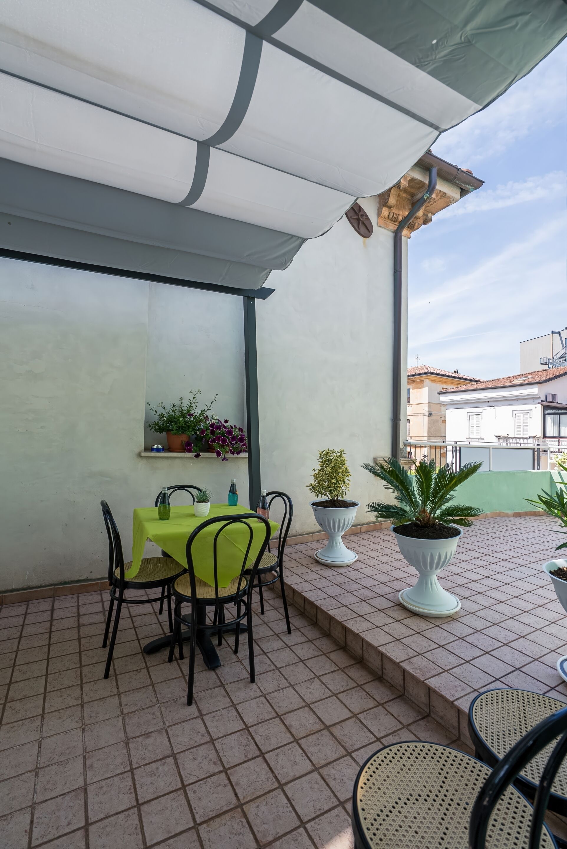 Um pátio coberto ao ar livre com mesa, cadeiras, plantas em vasos e vista para edifícios ao fundo.