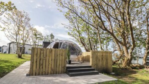 Uma trilha leva a uma estrutura em cúpula transparente, cercada por cercas de madeira e árvores.