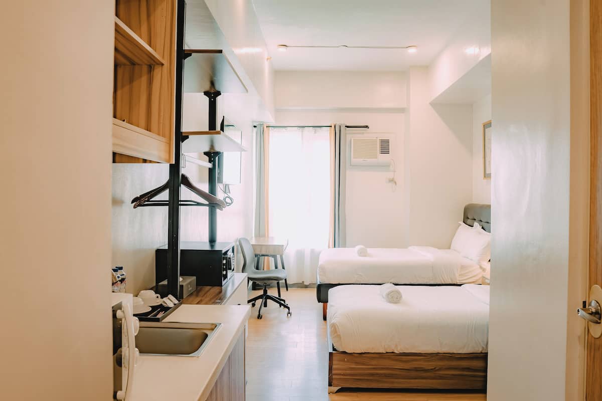 Quarto de hotel com duas camas, uma escrivaninha com cadeira, uma mesinha e um guarda-roupa.