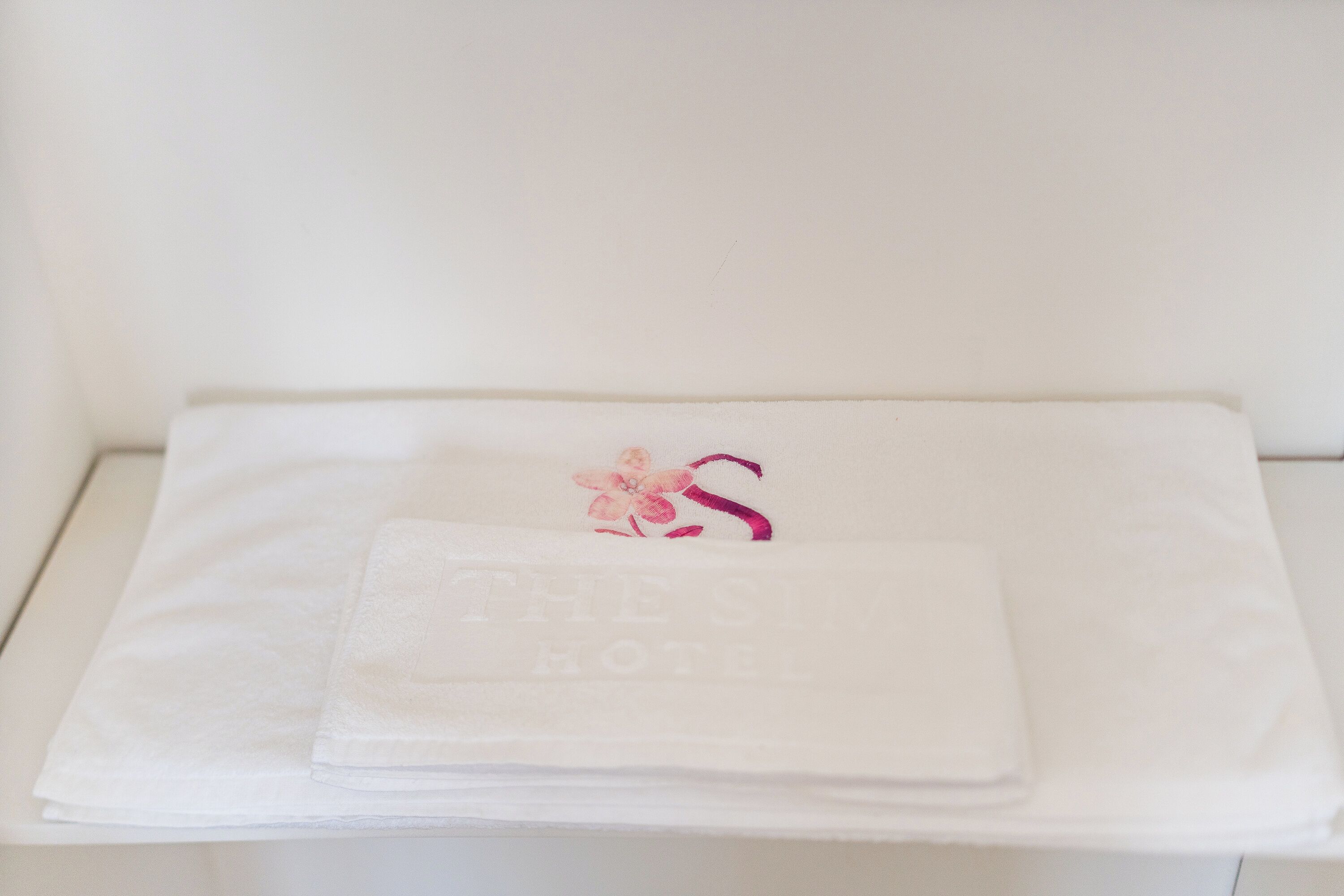 Toalha branca dobrada, com bordado de texto e uma flor, sobre uma superfície branca, provavelmente num quarto de hotel.