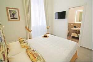Quarto com uma cama, televisão fixada na parede e banheiro visível por uma porta aberta.