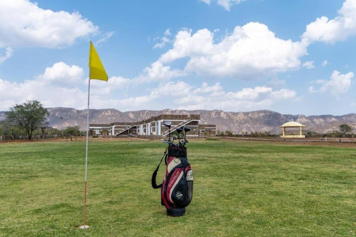 Uma mala de golfe em um campo verde com uma bandeira, um edifício e montanhas ao fundo.