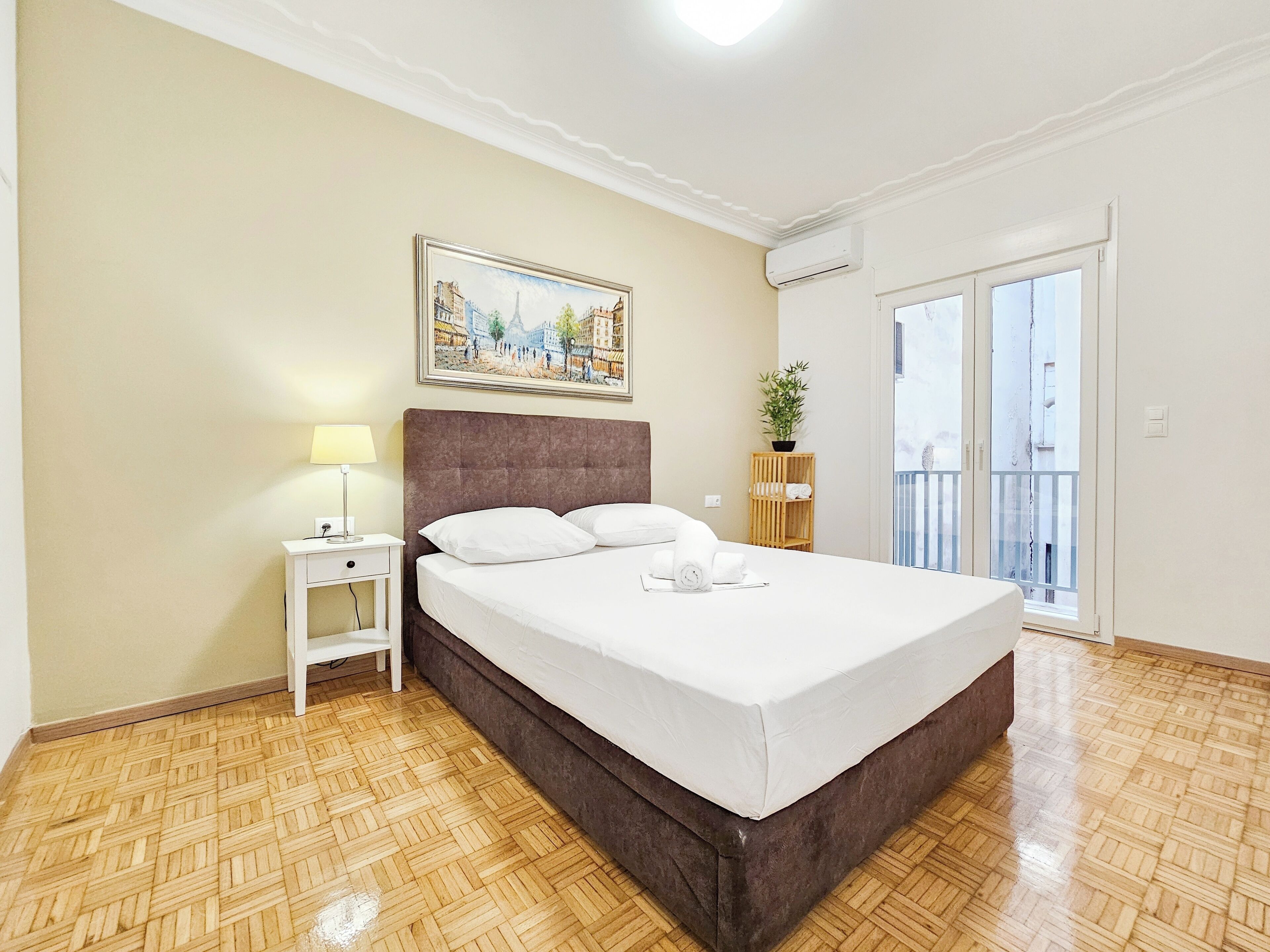 Quarto de hotel com uma cama grande, mesa de cabeceira com luminária, pintura de paisagem urbana, planta em vaso e varanda com grade.