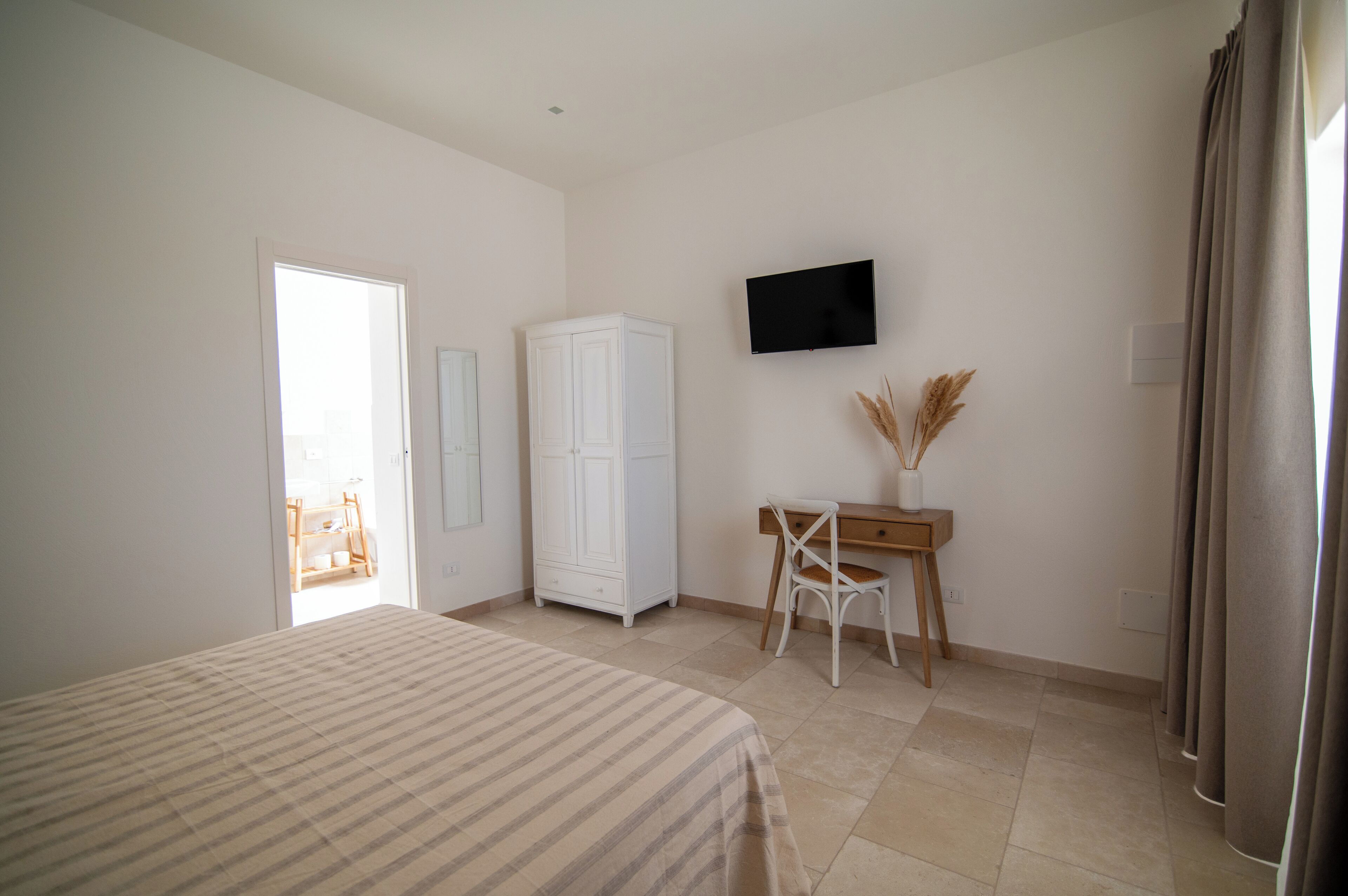 Quarto com cama, escrivaninha, cadeira, guarda-roupa, televisão e janela com cortinas.