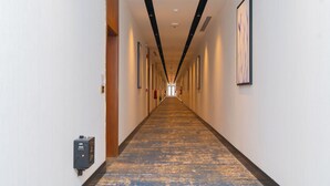 Um corredor de hotel comprido e estreito com carpete estampado, obras de arte emolduradas nas paredes e um alarme de incêndio à esquerda.