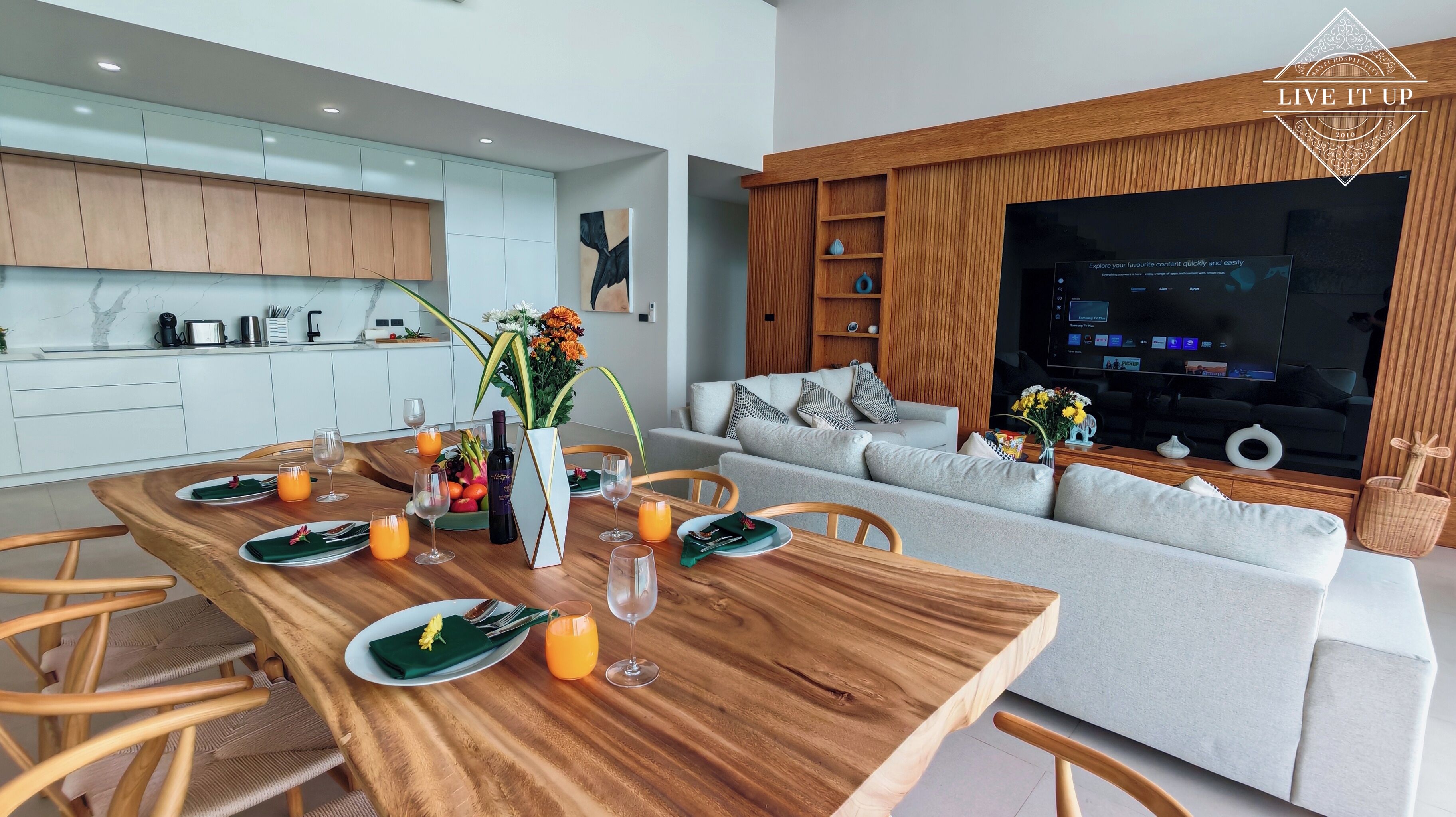 Quarto de hotel moderno com uma mesa de jantar de madeira posta para o café da manhã, um sofá cinza e uma grande tela de TV.
