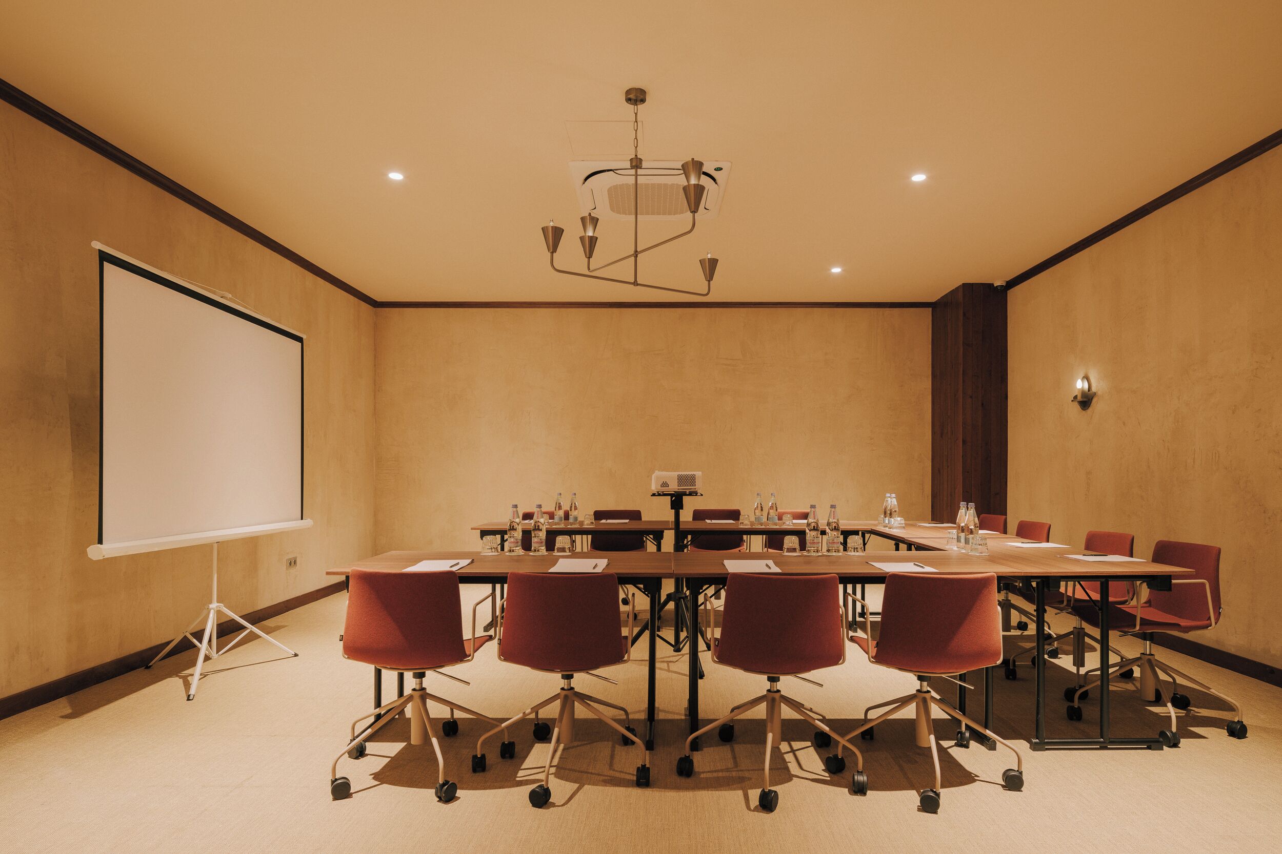 Uma sala de conferências com uma mesa grande, cadeiras vermelhas, uma tela de projetor e um lustre.