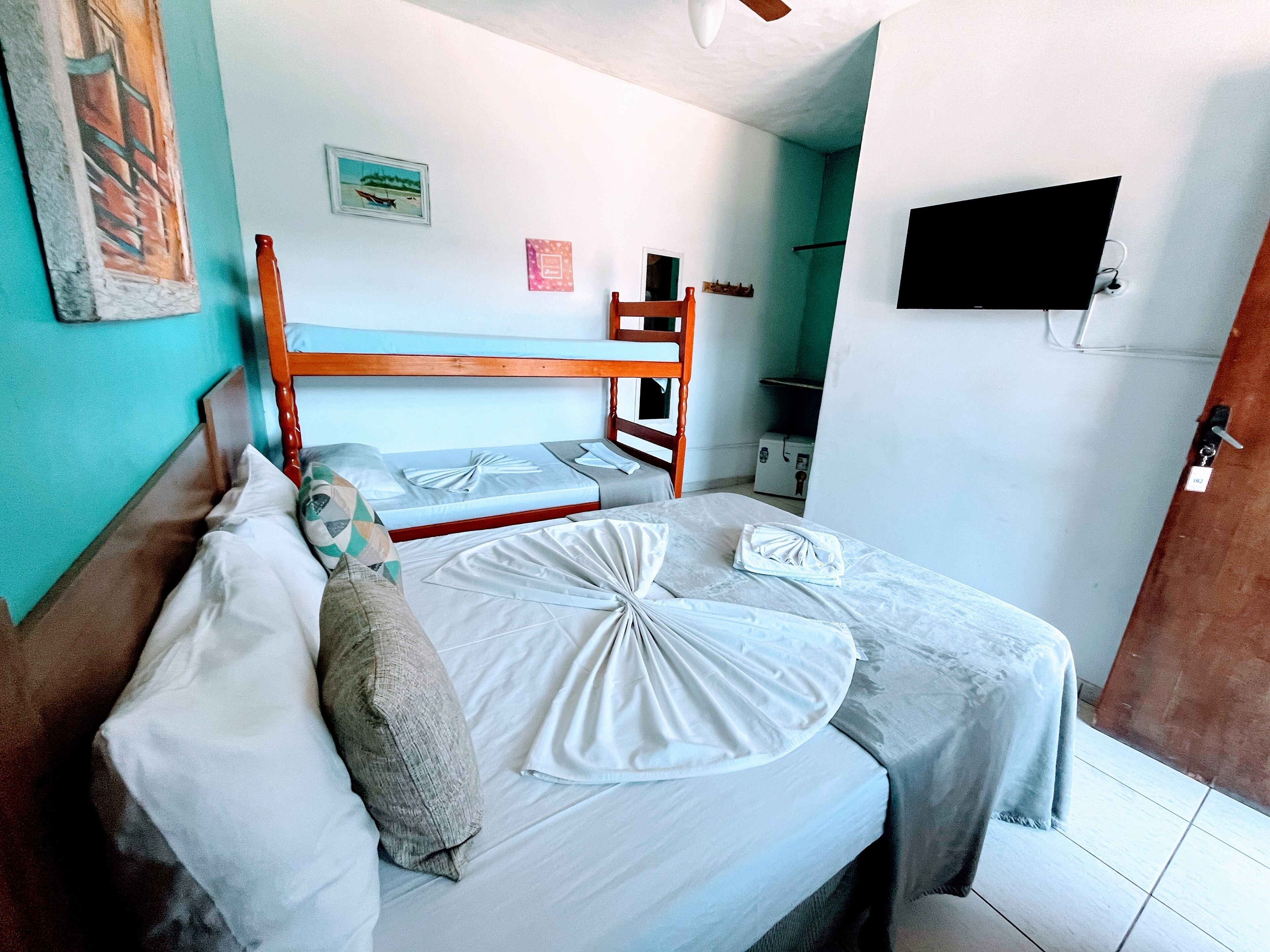 Uma cama de solteiro com cabeceira de madeira, uma mesinha de cabeceira e uma televisão fixada na parede.