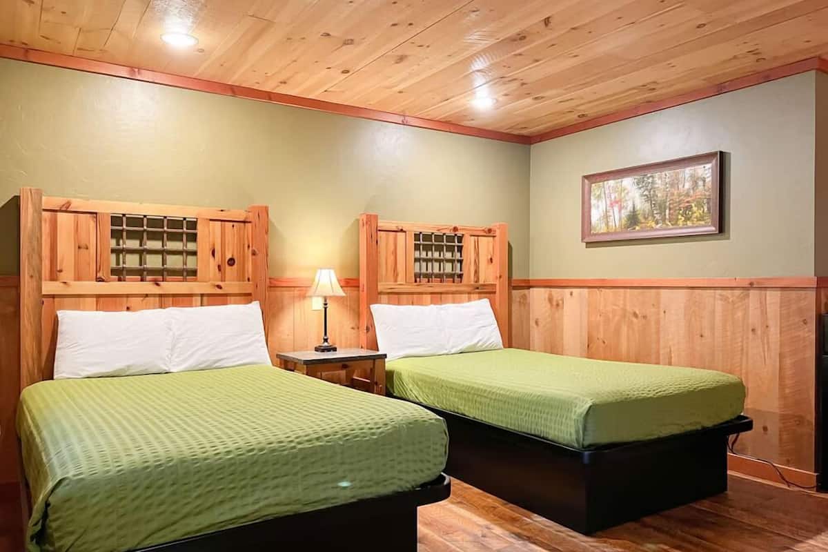 Quarto com duas camas, cabeceiras de madeira, uma pequena mesa com abajur e um quadro pendurado na parede.