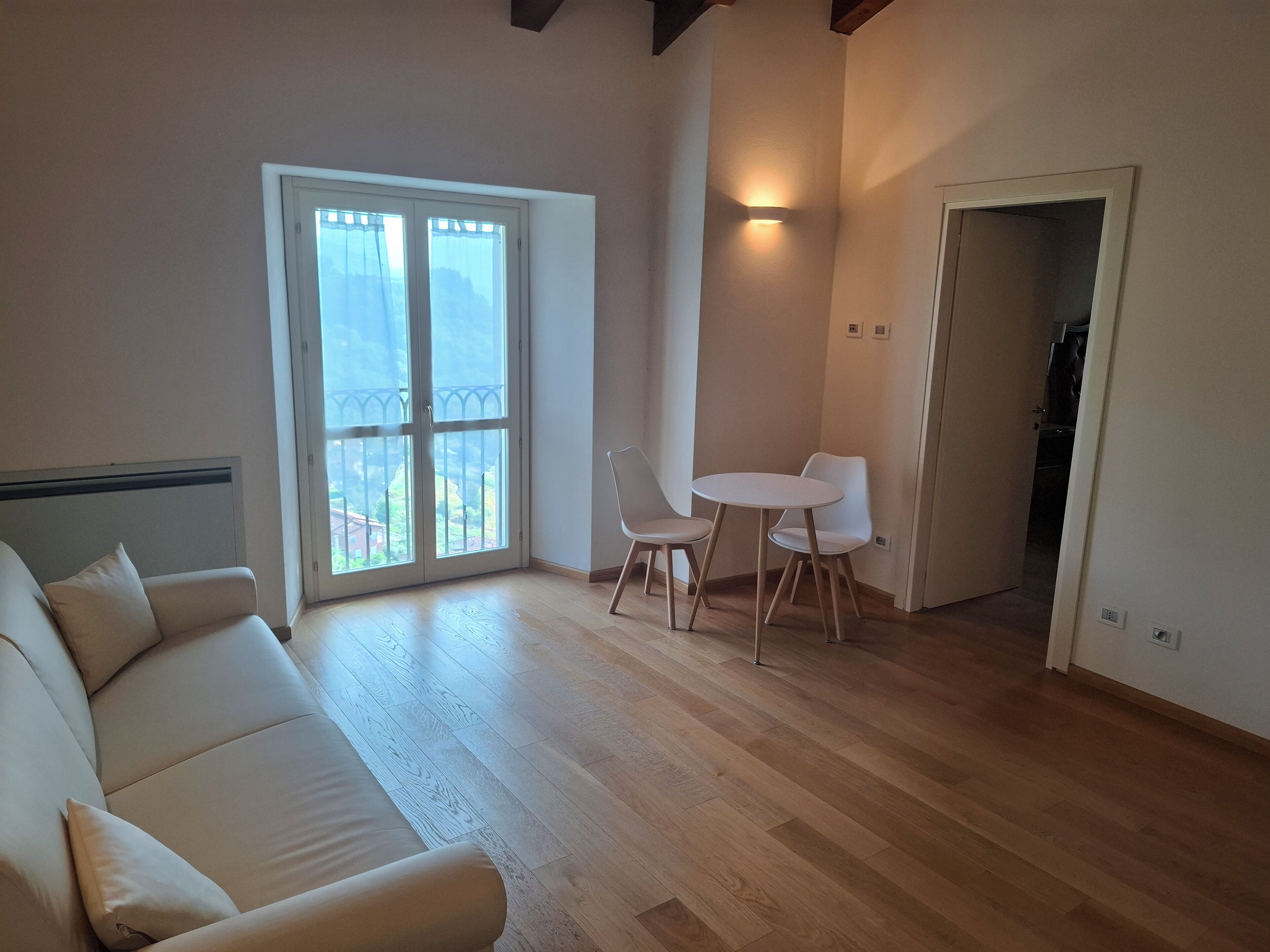 Uma sala ampla com uma grande porta de vidro deslizante, uma mesa redonda branca com duas cadeiras e um sofá bege.