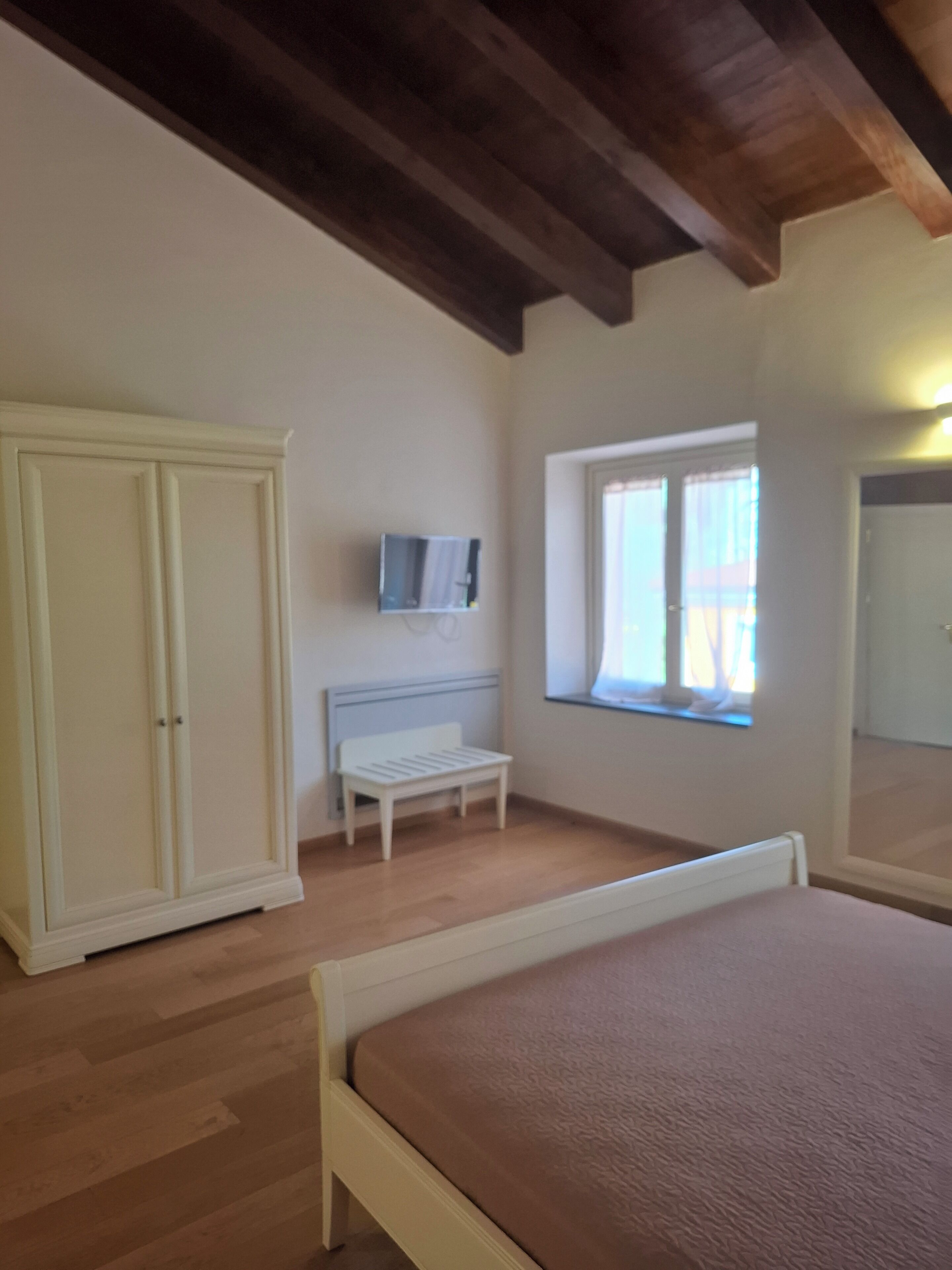 Um quarto com uma cama, um guarda-roupa, um banco, uma janela e uma televisão na parede.