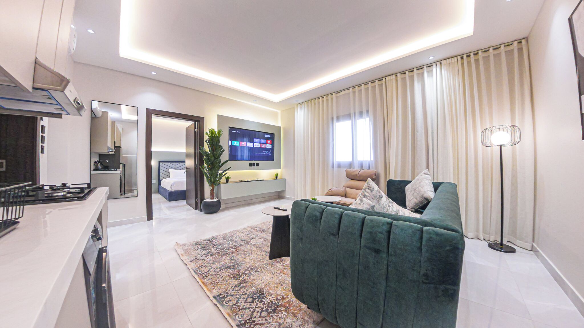 Sala de estar moderna com um sofá verde, uma televisão de tela plana e uma área de cozinha visível através de uma porta aberta.