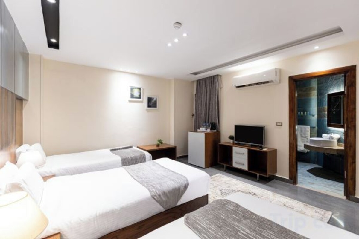 Quarto de hotel moderno com duas camas, uma televisão, uma escrivaninha e um banheiro visível através de uma porta aberta.