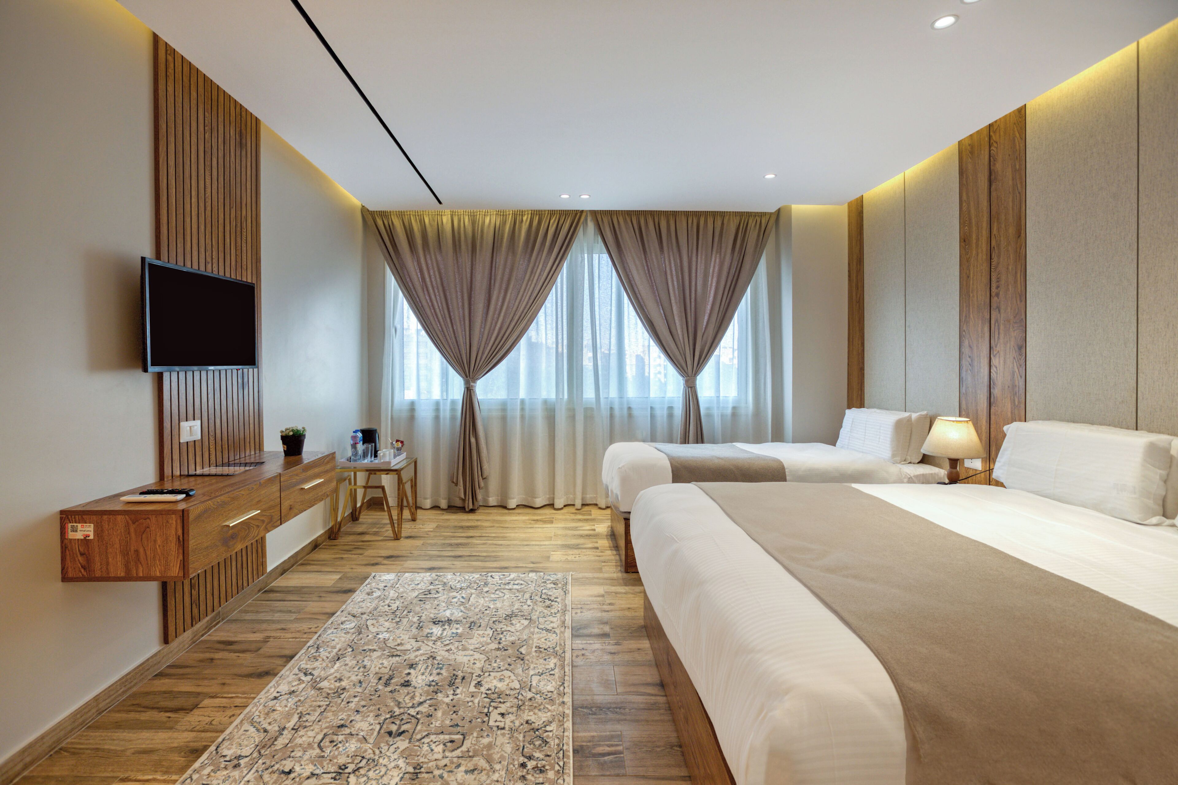 Quarto de hotel moderno com uma cama grande, uma televisão fixada na parede, uma escrivaninha com abajur e vista para a cidade através de amplas janelas.