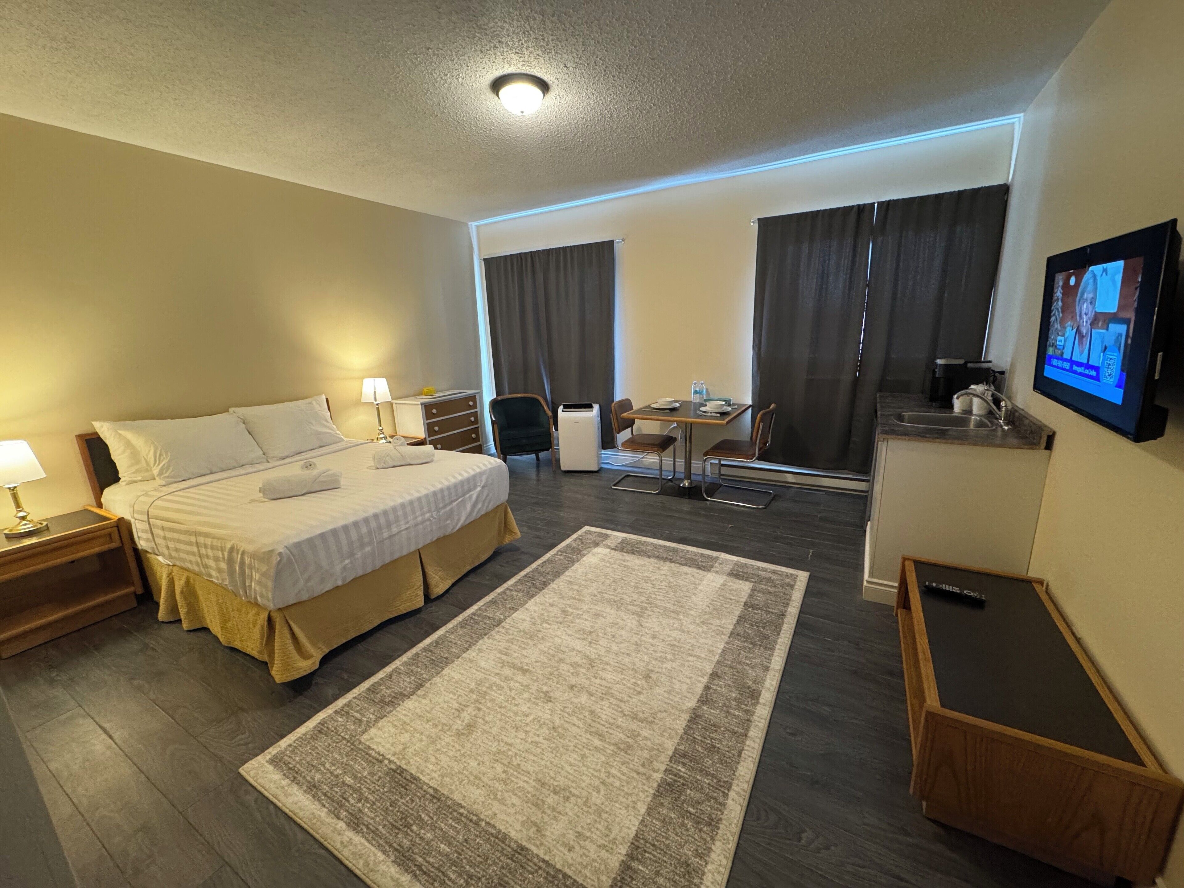 Quarto de hotel com cama, mesas de cabeceira, escrivaninha e cadeiras, televisão e uma pequena cozinha compacta.